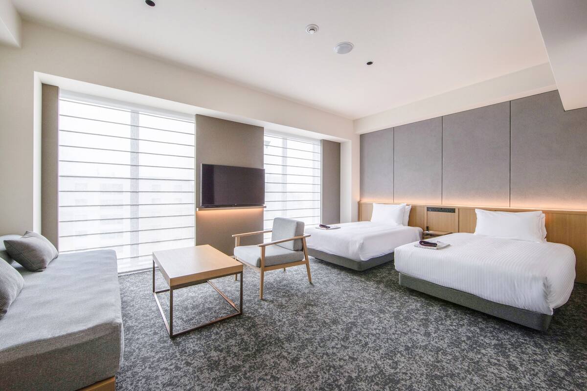 Quarto de hotel com duas camas, um sofá, uma mesinha e uma televisão.