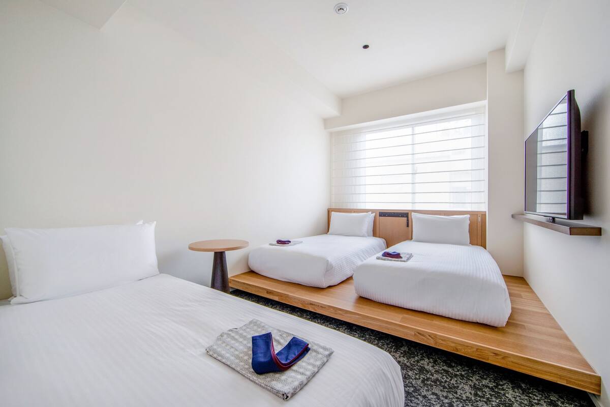 Quarto de hotel com duas camas, uma TV, uma mesinha e uma janela com persianas.