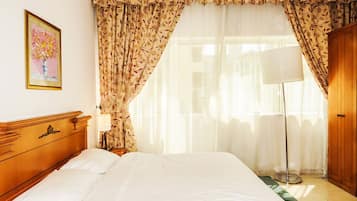 Quarto de hotel com uma cama grande, cabeceira de madeira, mesa de cabeceira com abajur e cortinas florais.