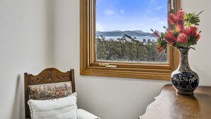 Quarto com vista para um lago e montanhas através de uma janela, uma cadeira de madeira com almofada estampada e um vaso com flores vermelhas sobre uma mesa de madeira.