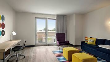 Quarto de hotel moderno com escrivaninha, cadeira de escritório, poltrona roxa, sofá azul, banquinho amarelo e tapete listrado colorido.