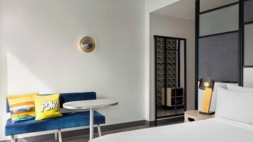 Quarto de hotel moderno com cama, mesa redonda, sofá azul com almofadas coloridas, luminária de parede e armário com portas deslizantes.
