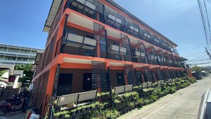Um edifício com vários andares e varandas, uma área de estacionamento com veículos estacionados e uma fileira de vasos de plantas ao longo da calçada.