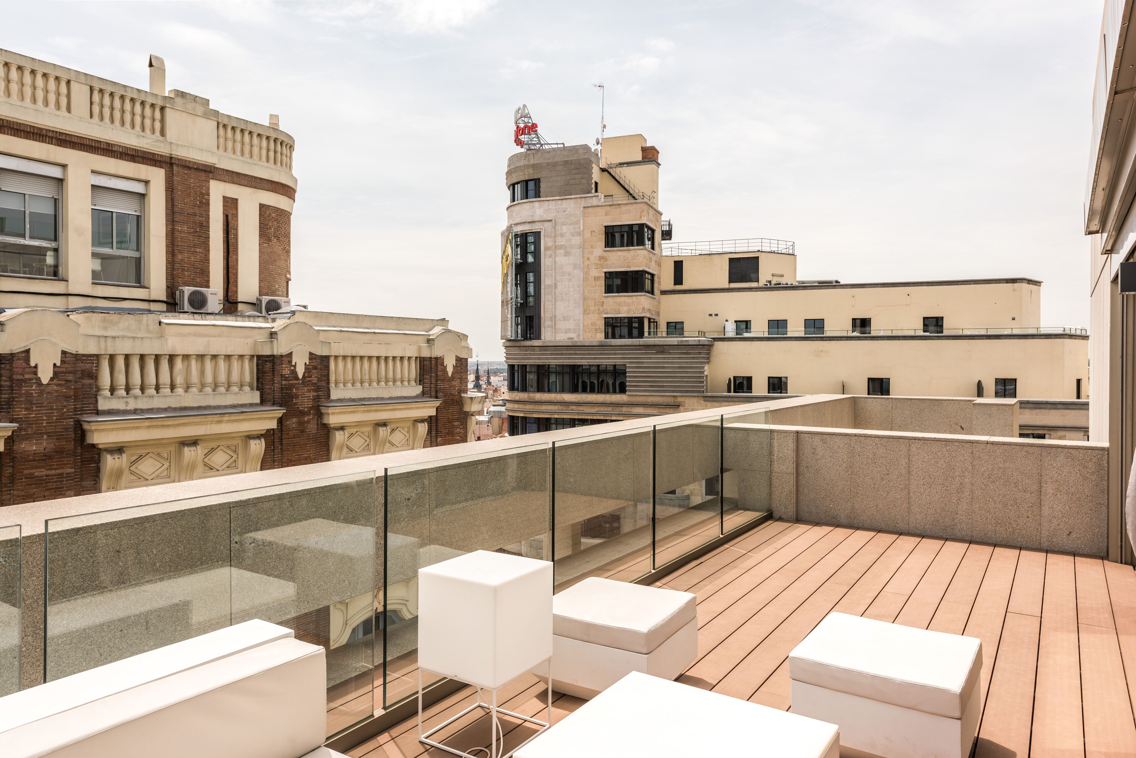 Terraço no telhado com guarda-corpos de vidro, assentos acolchoados brancos e vista para edifícios ao redor.