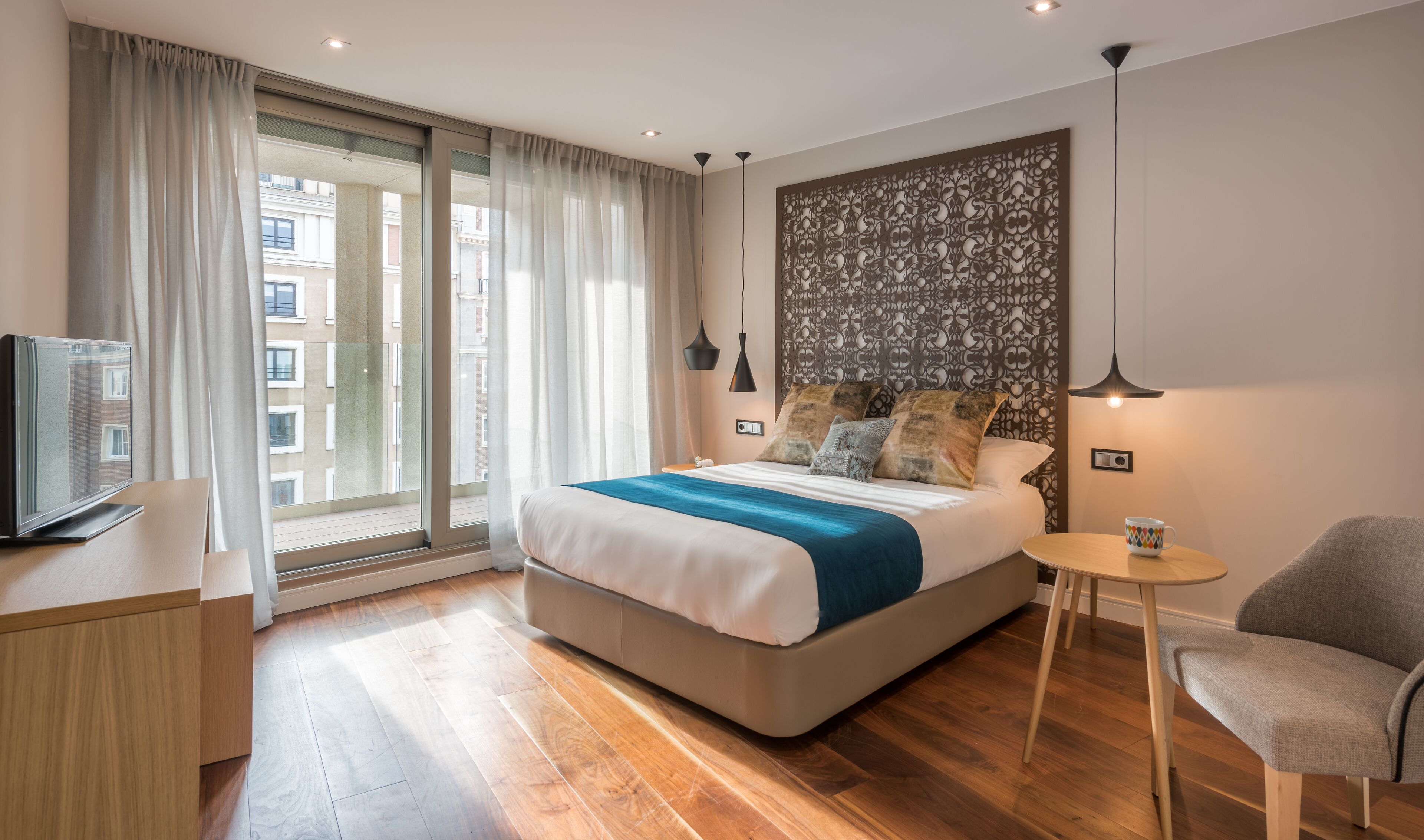 Quarto de hotel moderno com uma cama grande, televisão, poltrona e uma mesa pequena.