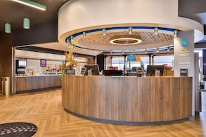 Uma área de recepção de hotel moderna, com um balcão de madeira, uma tela digital e um balcão exibindo comodidades.
