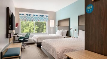 Quarto de hotel com duas camas, uma escrivaninha, uma cadeira e uma grande janela com vista para o verde.