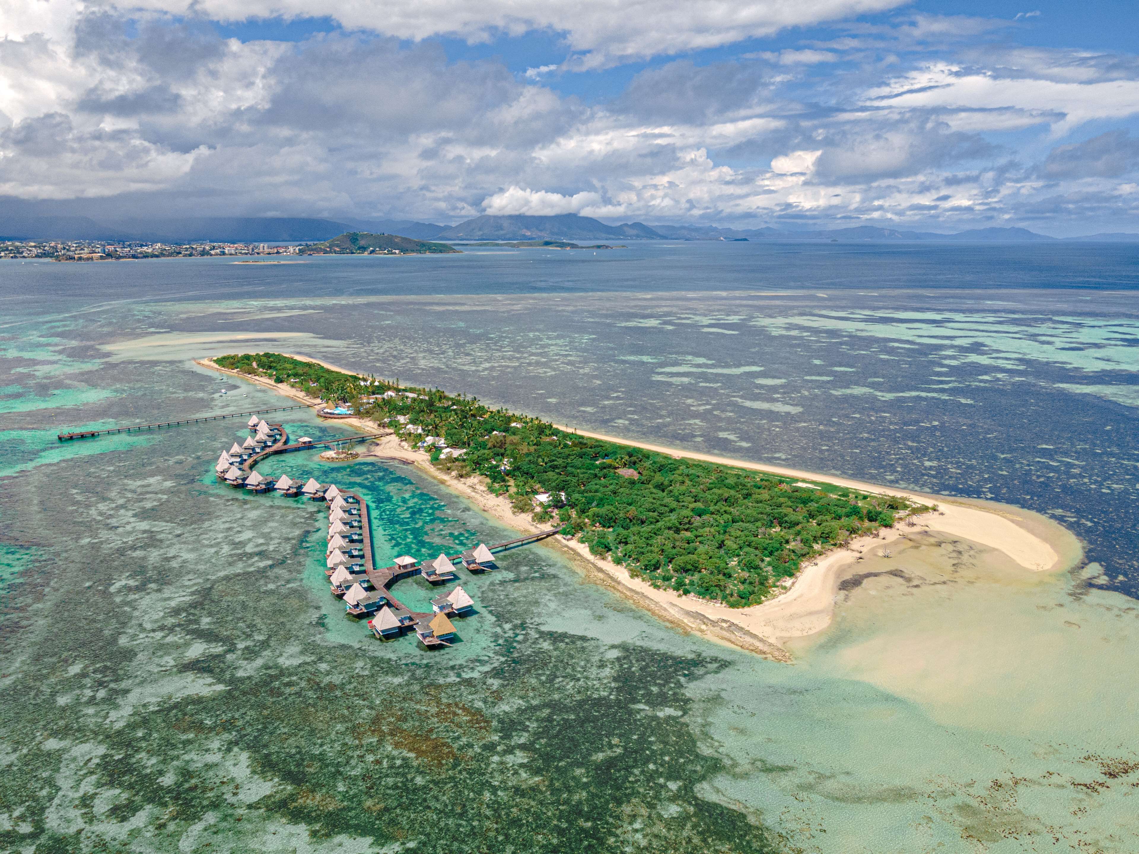 Vista aérea de uma ilha resort com bangalôs sobre a água, ligados por uma ponte.