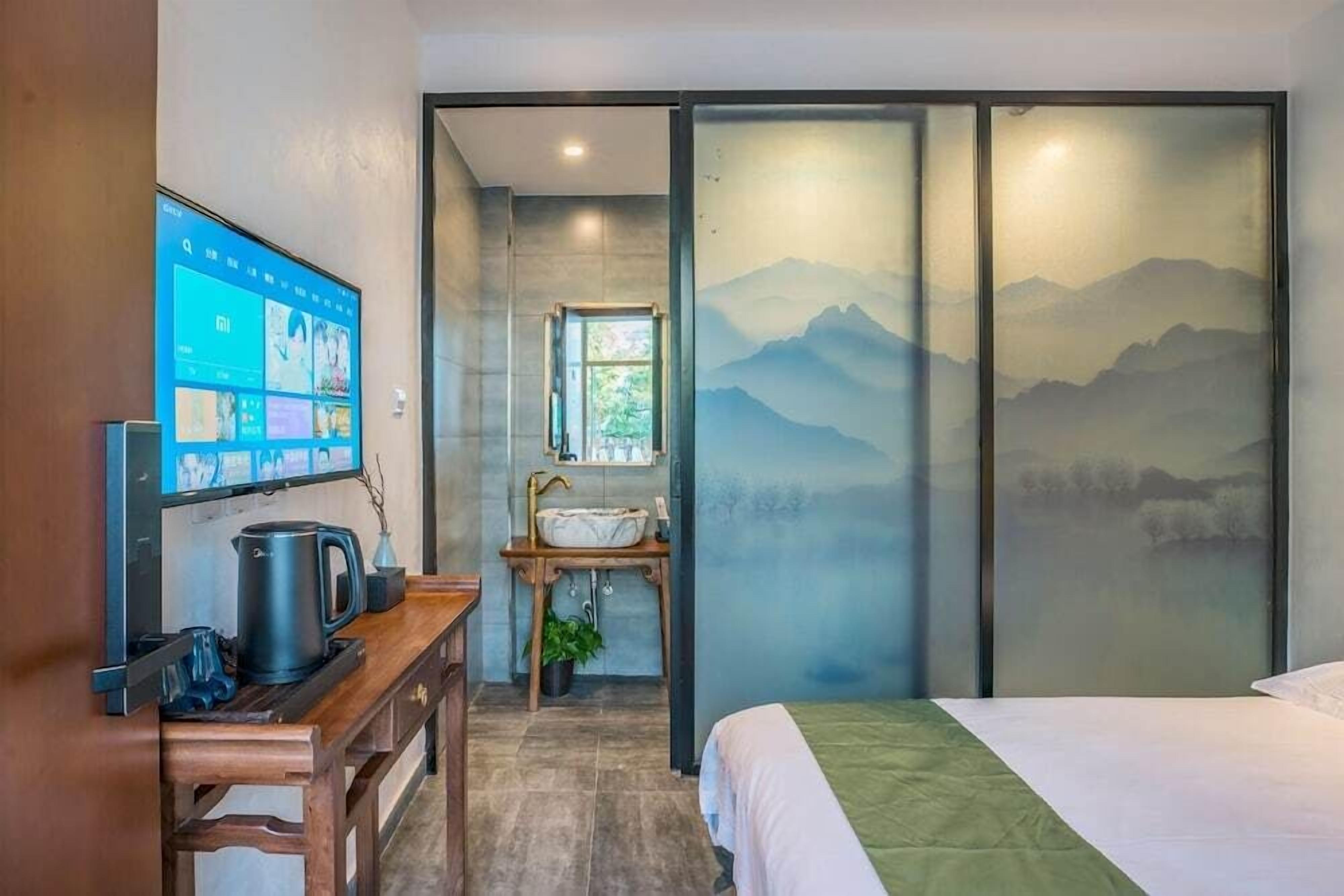 Quarto de hotel com cama, escrivaninha com chaleira, televisão exibindo um cardápio, janela e porta deslizante com vista para a montanha.