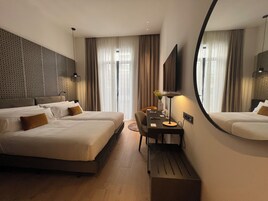 Quarto de hotel moderno com duas camas, uma escrivaninha com candeeiro e um espelho grande.