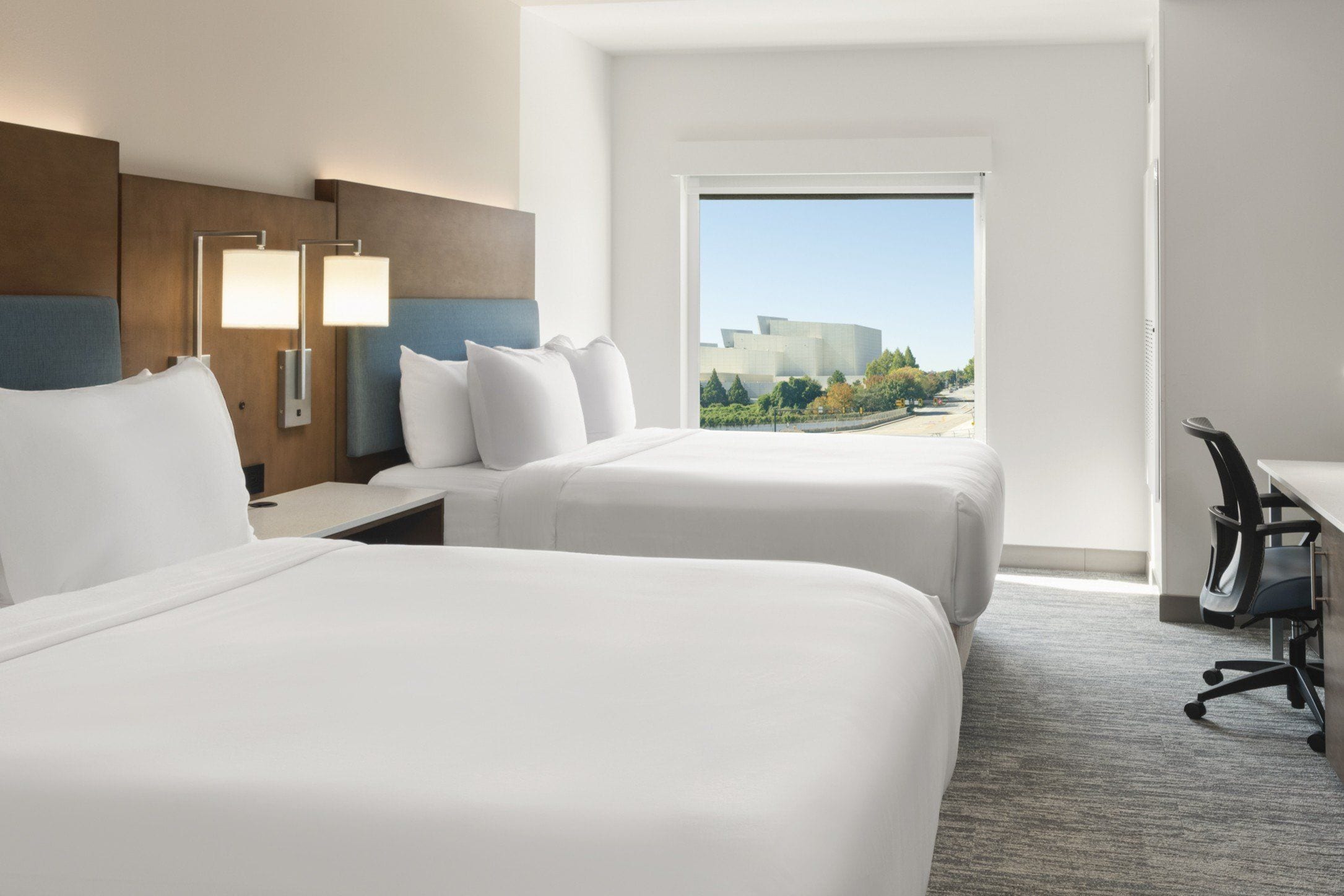 Quarto de hotel com duas camas, uma escrivaninha e uma cadeira. Há uma janela com vista para edifícios e árvores.