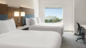 Quarto de hotel com duas camas, uma escrivaninha e uma cadeira. Há uma janela com vista para edifícios e árvores.
