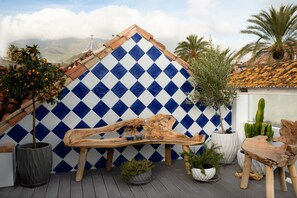 Um banco de madeira com design rústico, rodeado por vasos de plantas e uma parede com padrão geométrico azul e branco.