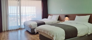 Quarto de hotel com duas camas, piso de madeira, uma janela grande com cortinas finas e uma luminária fixada na parede.