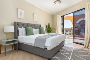 Quarto moderno com uma cama grande, mesa de cabeceira, abajur e vista para a cidade através de uma varanda.