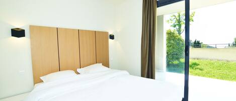 Quarto moderno com uma cama grande, cabeceira de madeira e porta de vidro deslizante para uma varanda com vista para o verde.