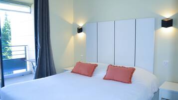 Quarto de hotel com uma cama grande, duas almofadas, uma mesinha de cabeceira e uma luminária fixada na parede.