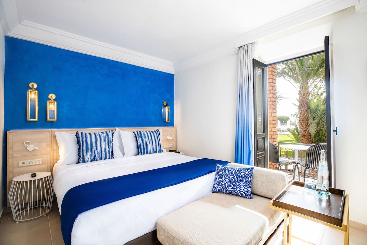 Quarto de hotel com uma cama grande, um sofá, uma mesinha e uma varanda com mobiliário de exterior.