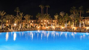 Uma área de piscina à noite com espreguiçadeiras e guarda-sóis, cercada por palmeiras e edifícios iluminados.