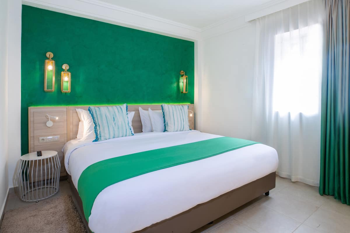 Uma cama bem arrumada com um cobertor verde, uma mesa de cabeceira com uma luminária e uma parede com duas arandelas.