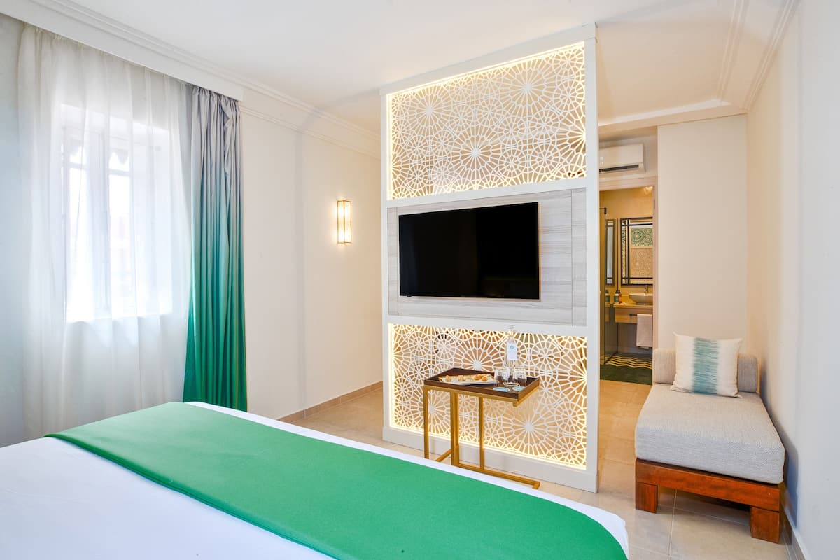 Quarto de hotel com cama, televisão fixada em painel decorativo, mesinha com bandeja e um banco com almofadas.
