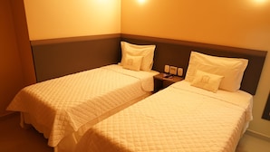 Quarto de hotel com duas camas, cada uma com colcha acolchoada e almofadas a combinar, mesa de cabeceira com abajur e iluminação embutida na parede.