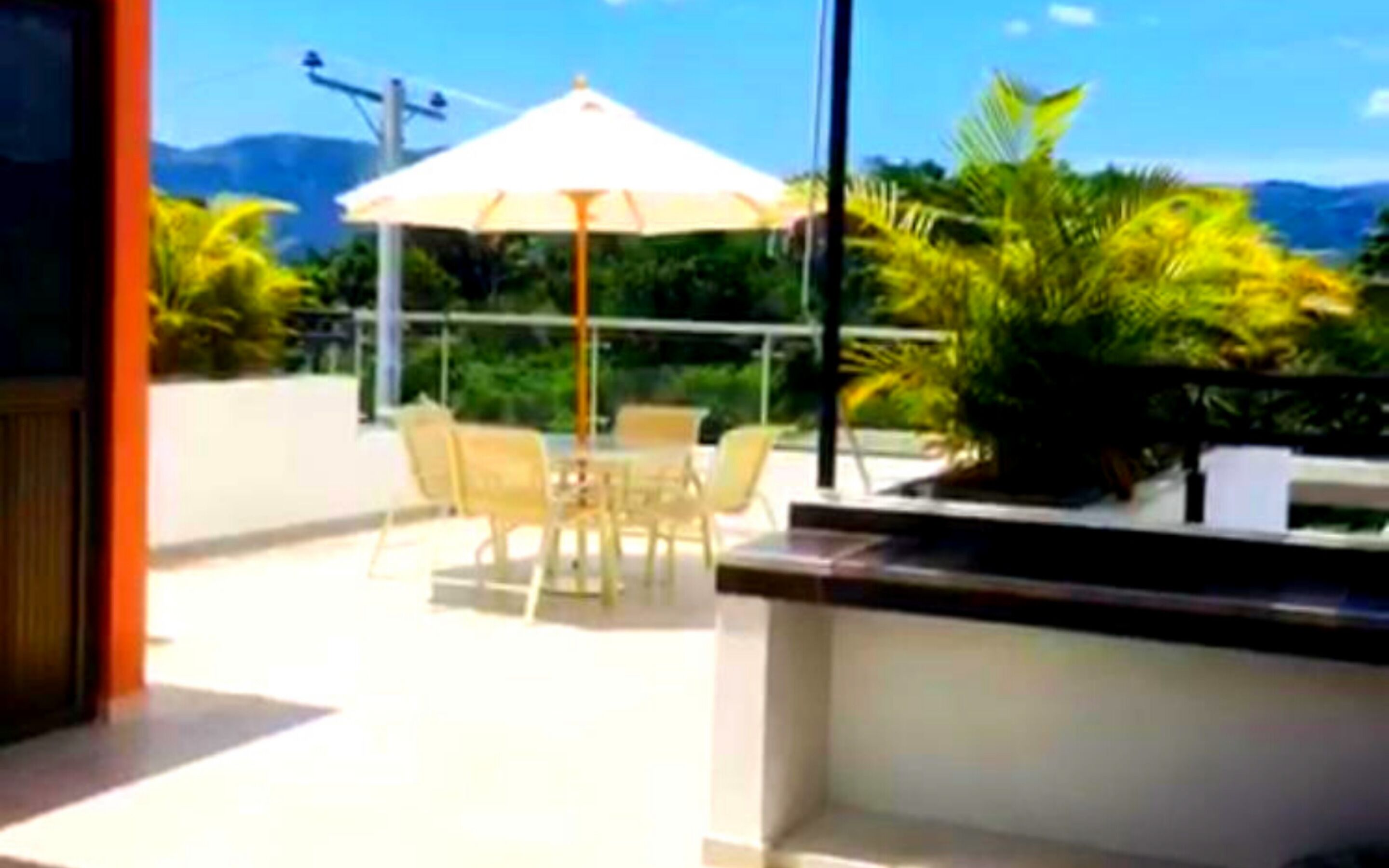 Um pátio externo com mesa, cadeiras, um guarda-sol branco e plantas em vasos.