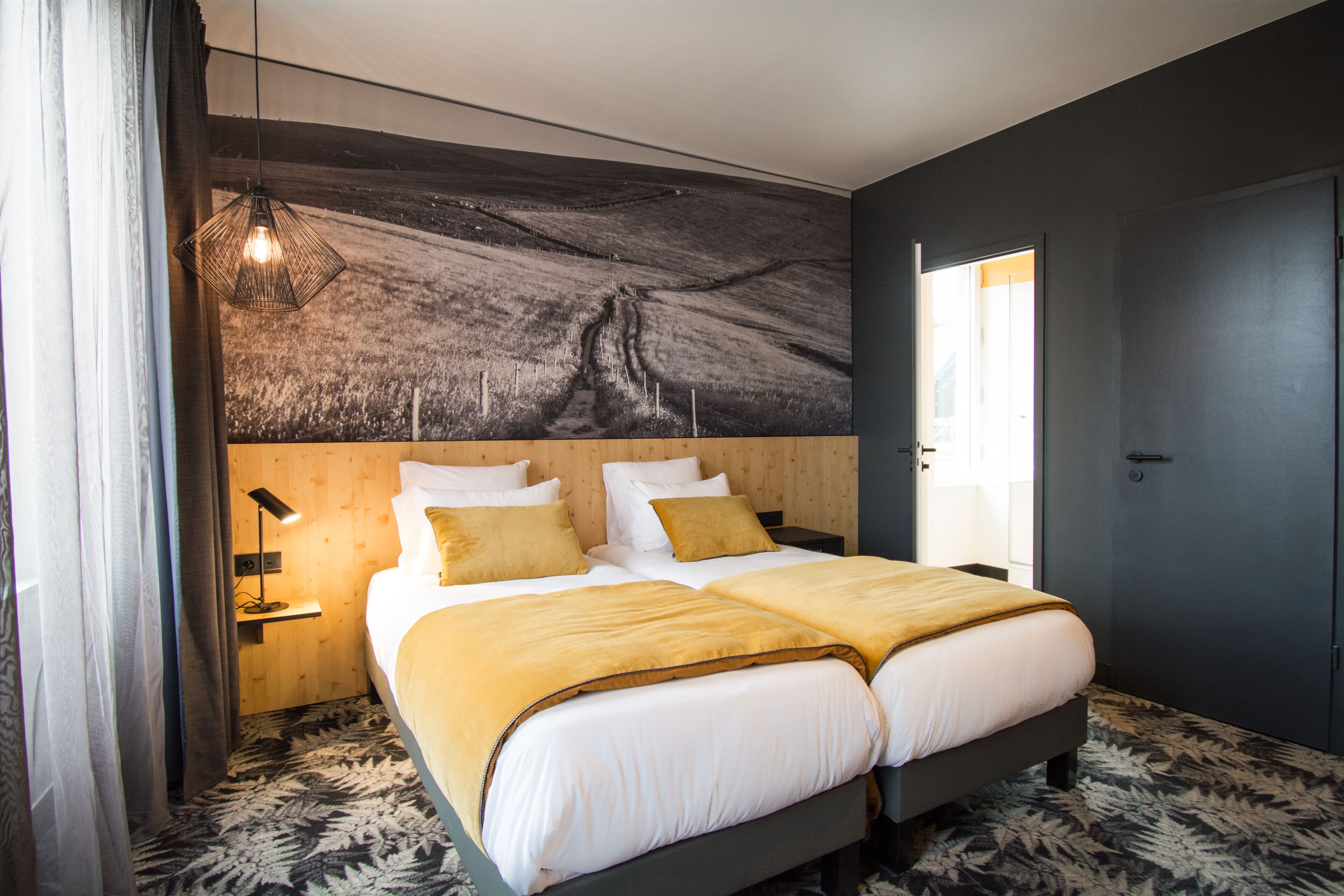Uma cama de casal com duas almofadas, uma mesa de cabeceira com abajur, um mural grande na parede e uma porta à direita.