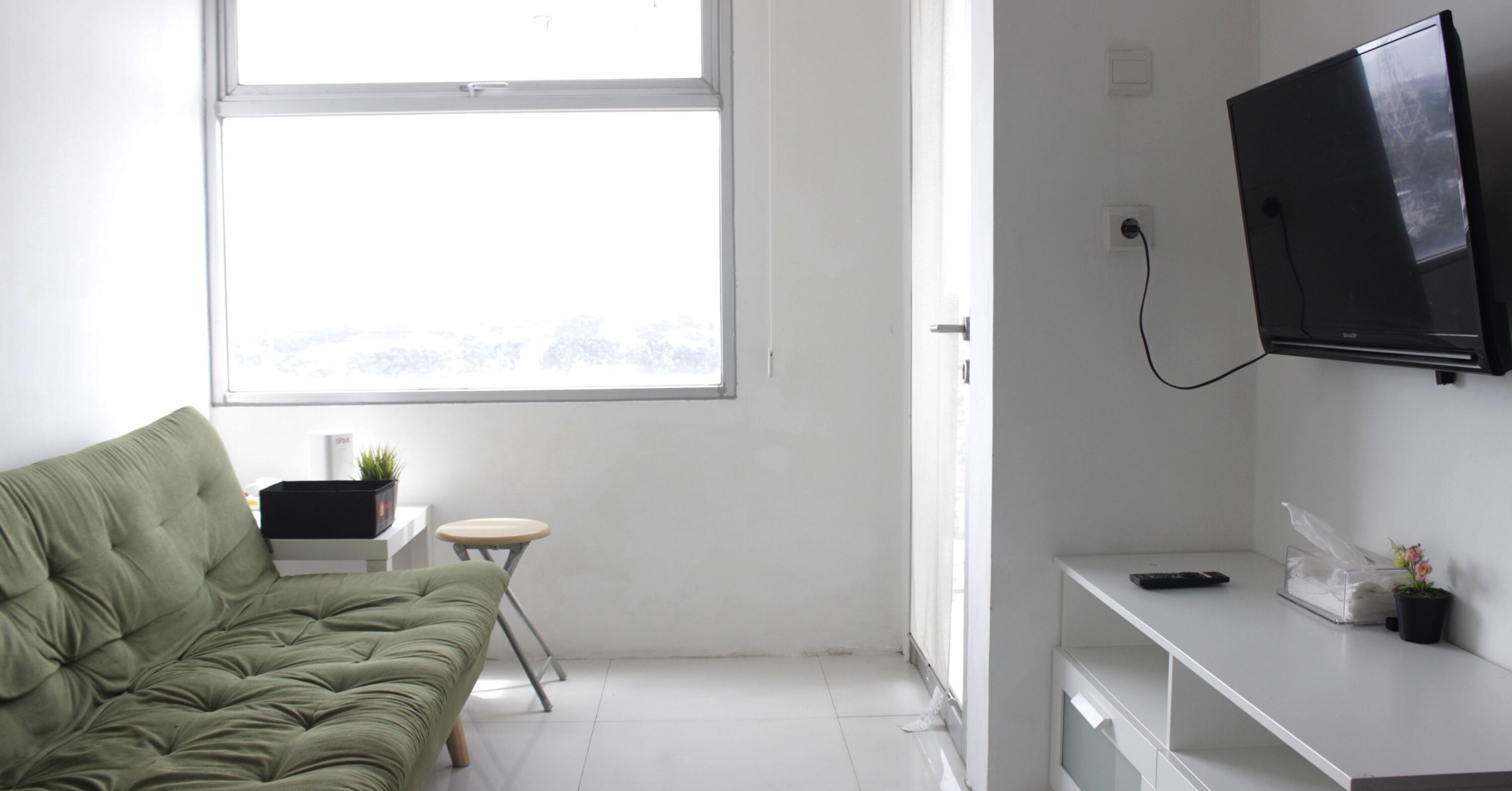 Quarto minimalista com um sofá verde, uma mesinha branca, uma televisão na parede e uma janela com vista.