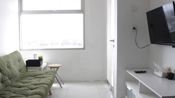 Quarto minimalista com um sofá verde, uma mesinha branca, uma televisão na parede e uma janela com vista.