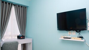 Quarto com escrivaninha, cadeira, televisão e uma estante com dispositivos eletrônicos.