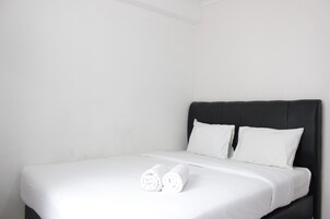 Cama bem arrumada com lençóis brancos e duas toalhas enroladas, cabeceira preta e paredes brancas.