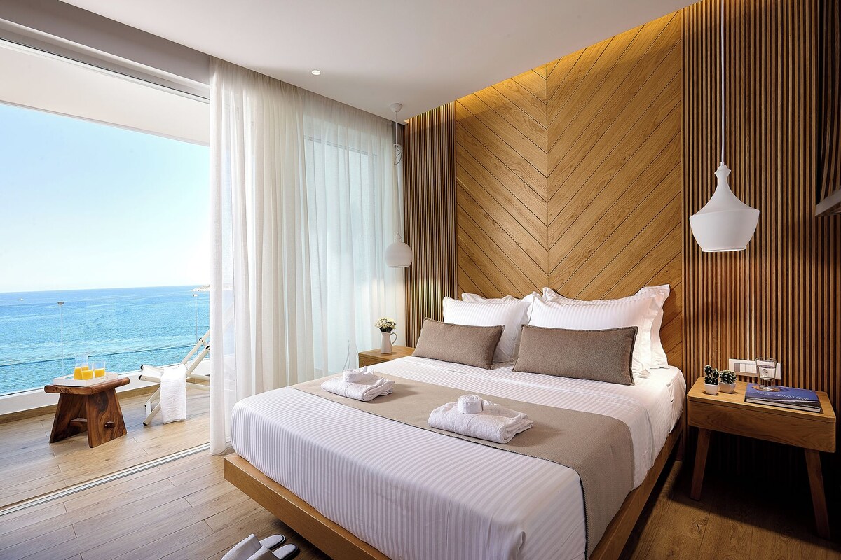 Quarto de hotel com uma cama grande, cabeceira de madeira, mesa de cabeceira com abajur e vista para o mar.