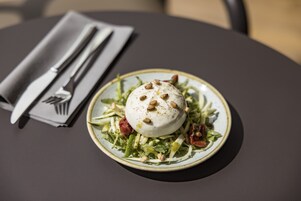 Prato de comida com um item esférico branco sobre uma base de folhas verdes e frutos secos, acompanhado de talheres e guardanapo sobre uma mesa redonda.