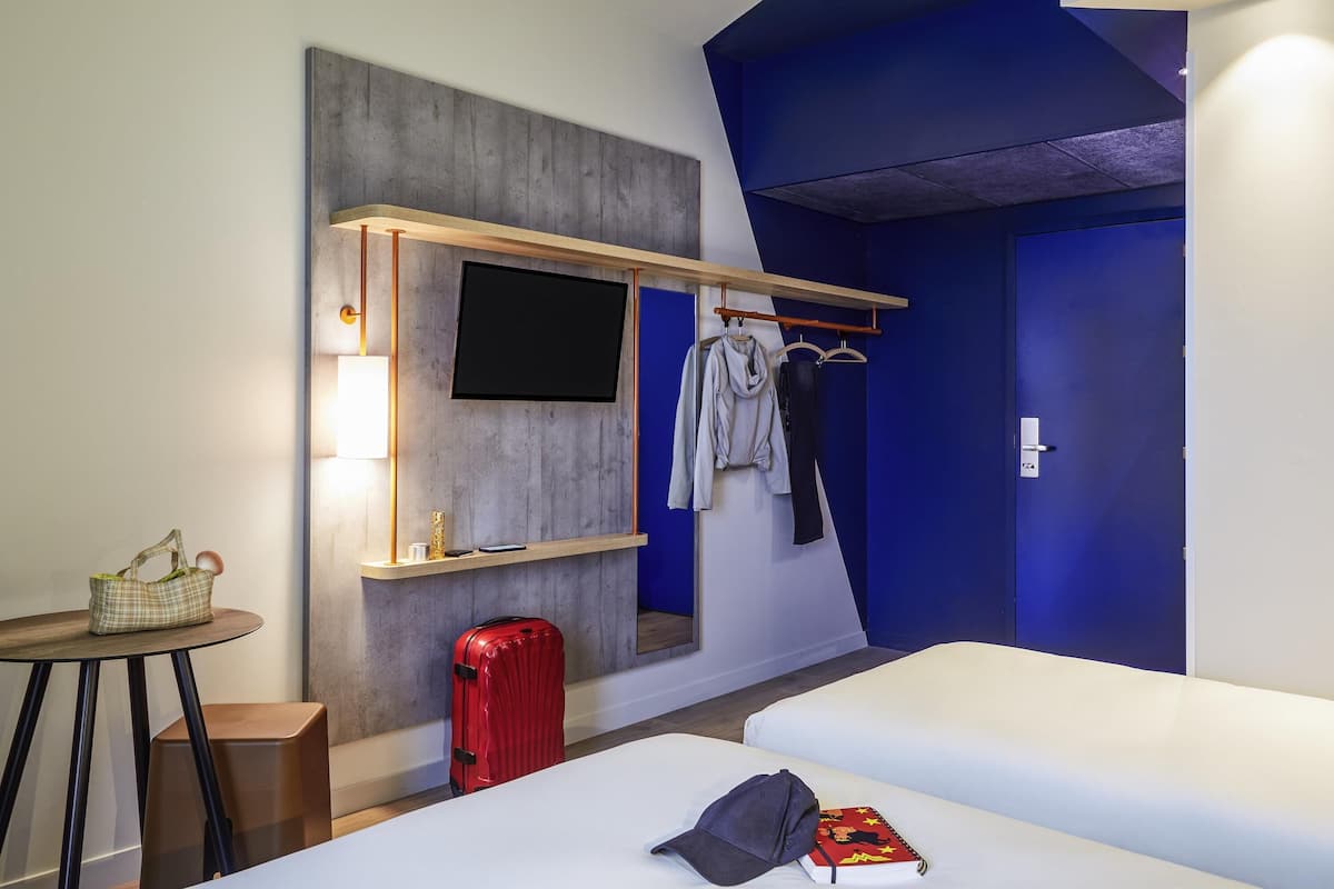 Quarto de hotel com uma cama, televisão na parede, cabideiro com roupas, mala, mesinha com abajur e bolsa.