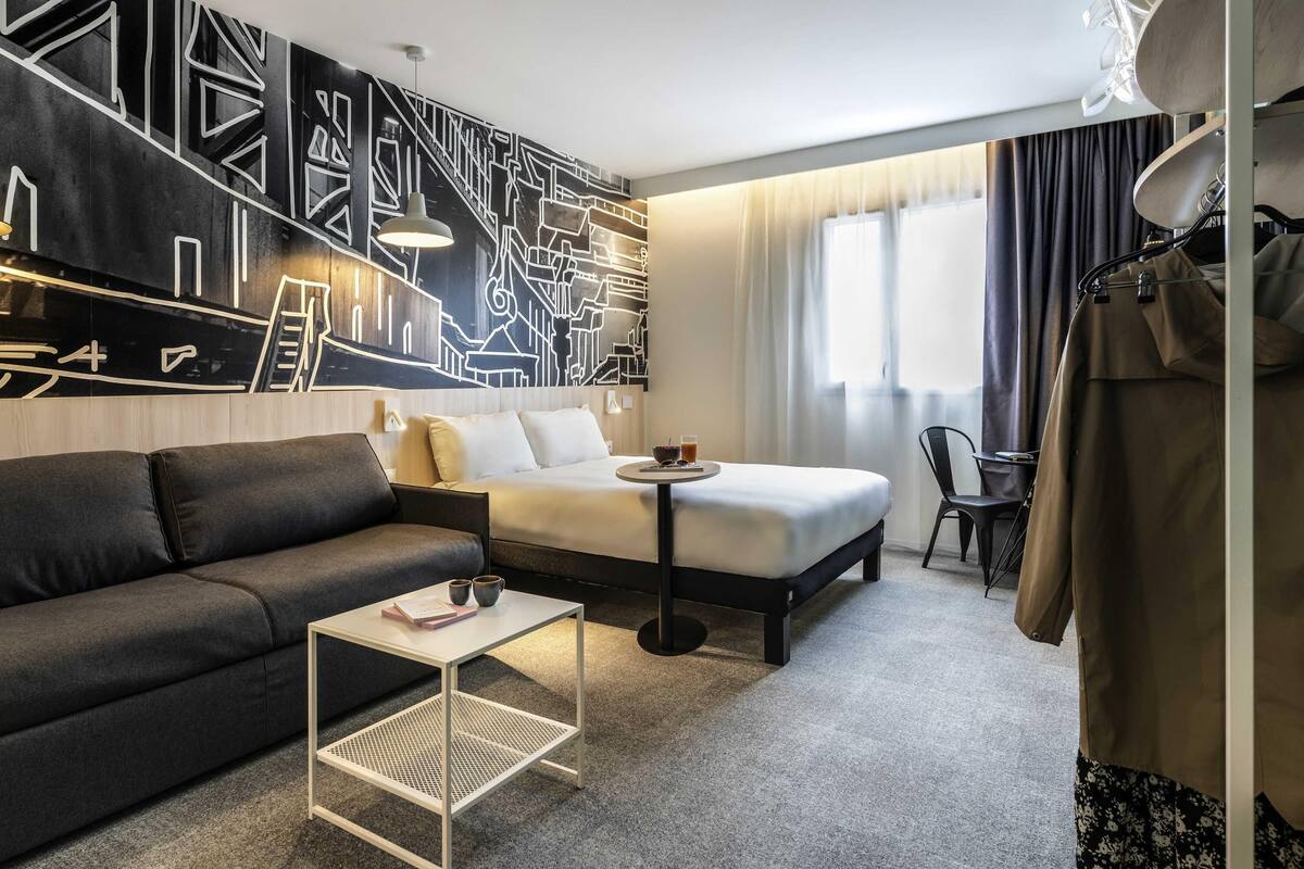 Quarto de hotel com cama, sofá, mesinha, cadeira, porta-casacos e um grande mural abstrato na parede.