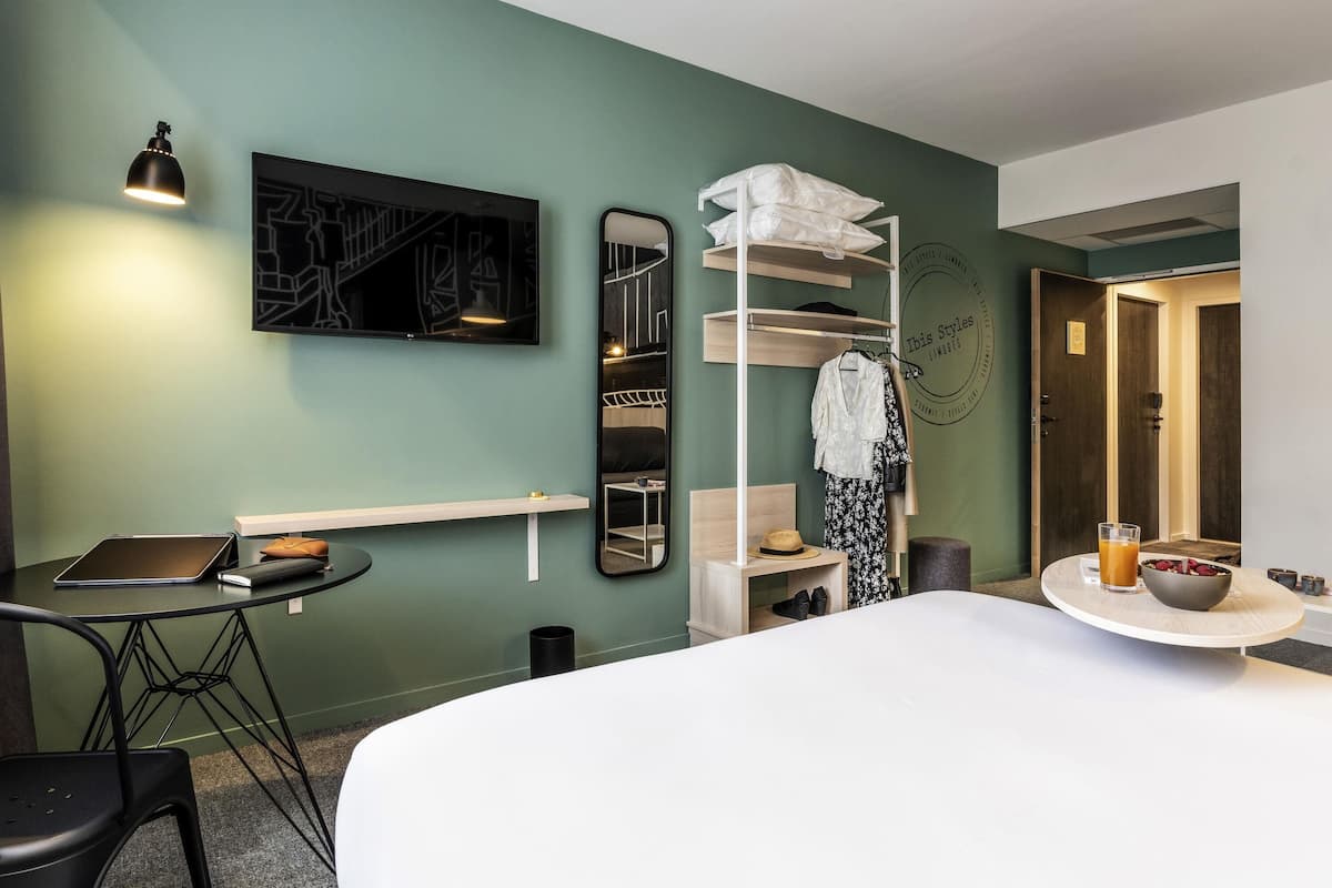Quarto de hotel moderno com cama, escrivaninha, cadeira, televisão, cabideiro e armário.