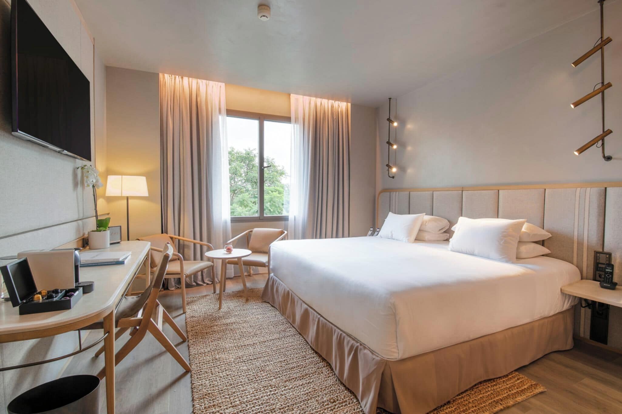 Quarto de hotel com cama, mesa de cabeceira, abajur, escrivaninha, cadeira e vista para o exterior.