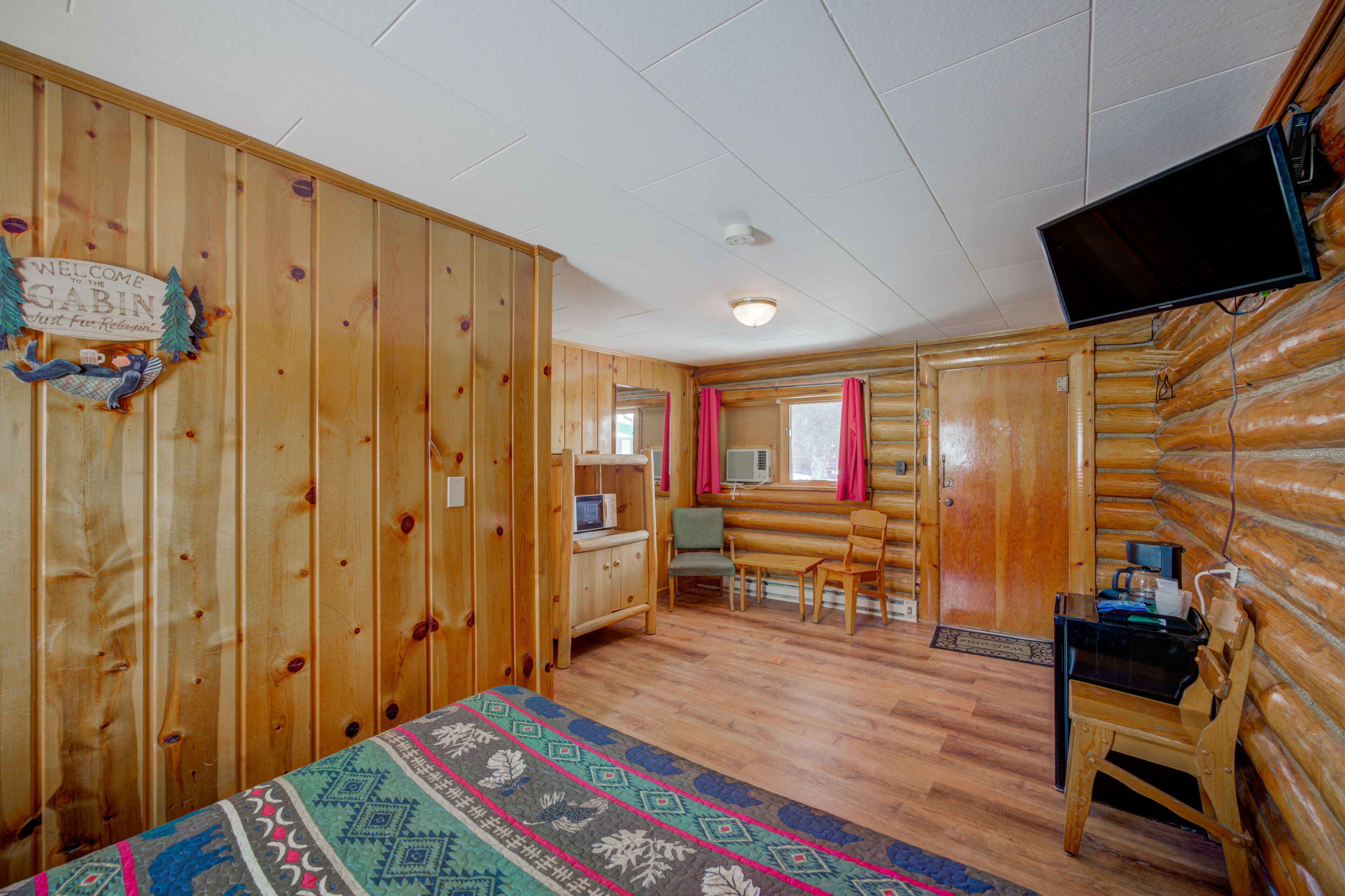 Quarto estilo cabana com paredes de madeira, uma cama com colcha estampada, uma escrivaninha com computador, uma televisão na parede e uma janela com cortinas cor-de-rosa.