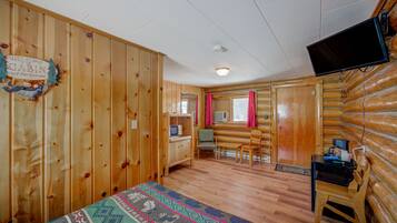 Quarto estilo cabana com paredes de madeira, uma cama com colcha estampada, uma escrivaninha com computador, uma televisão na parede e uma janela com cortinas cor-de-rosa.