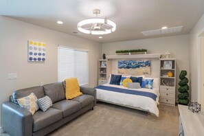 Um quarto com cama, sofá cinza, ventilador de teto, janela com persianas, um quadro e uma estante com objetos decorativos.