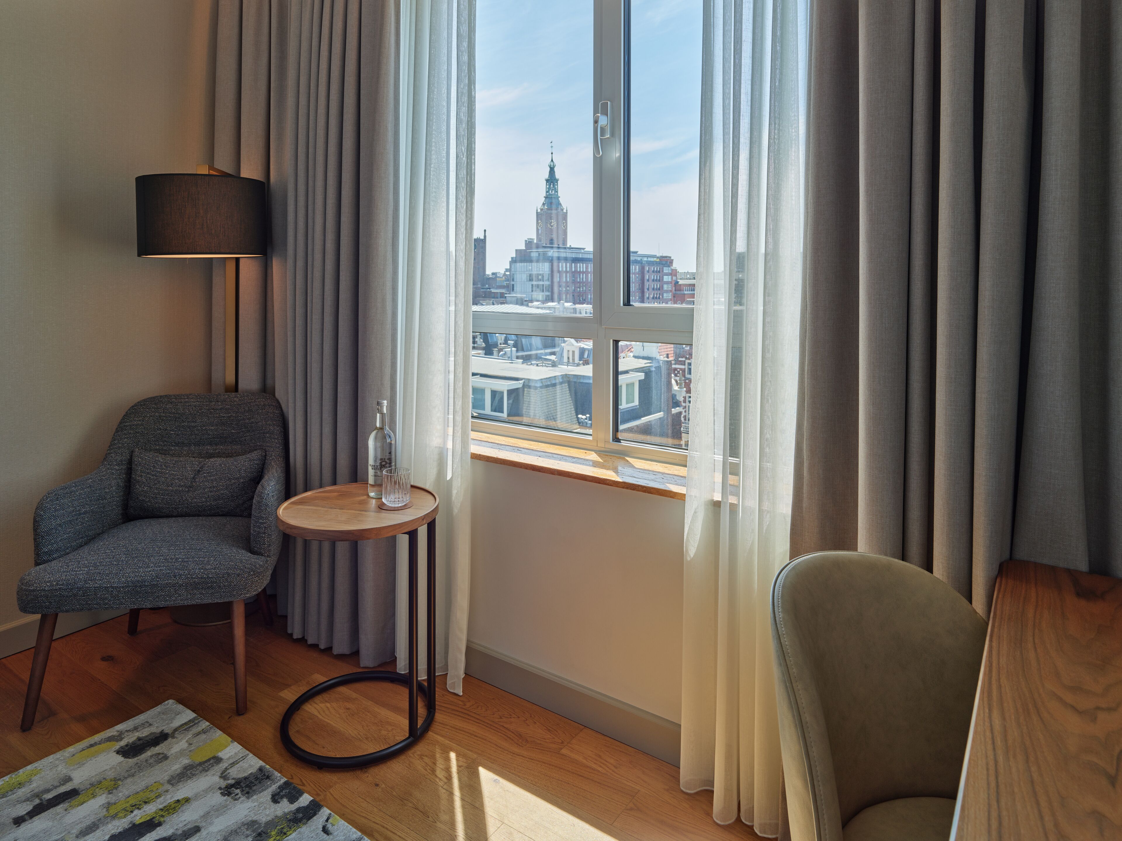 Quarto de hotel moderno com vista para a paisagem urbana, uma poltrona cinza, uma mesinha redonda com uma garrafa e um copo, e um abajur.