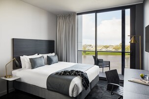 Quarto de hotel moderno com uma cama grande, uma escrivaninha com cadeira e vista para a cidade através de uma grande janela.