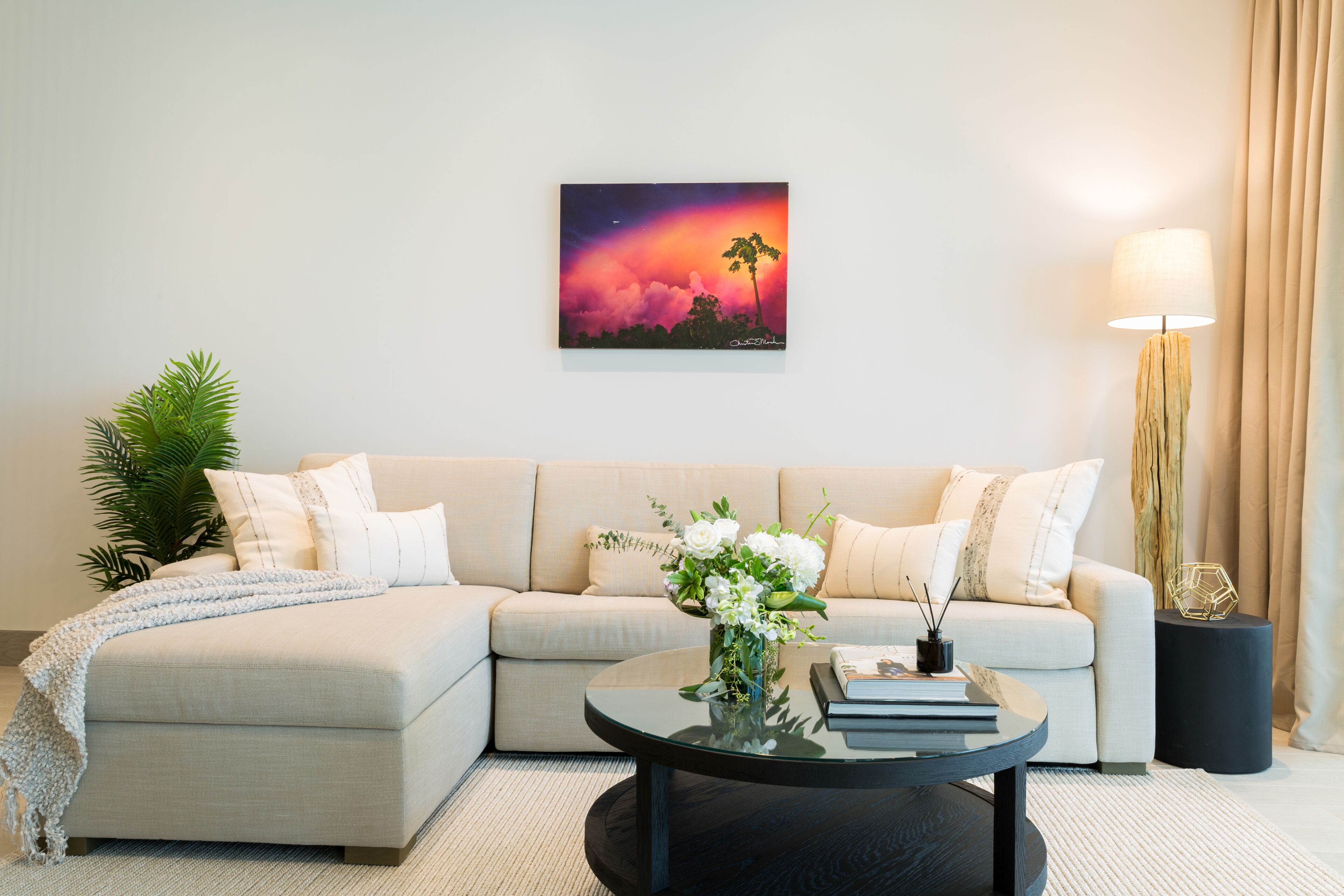 Uma sala de estar moderna com um sofá bege, uma mesa de centro redonda com um vaso de flores, e um quadro com um pôr do sol na parede.