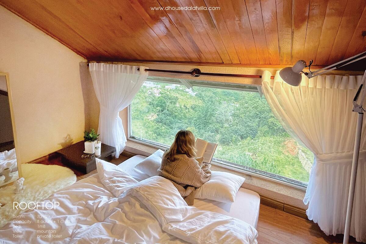 Quarto com cama, janela com cortinas, mesa de cabeceira com abajur e vista para o verde lá fora.