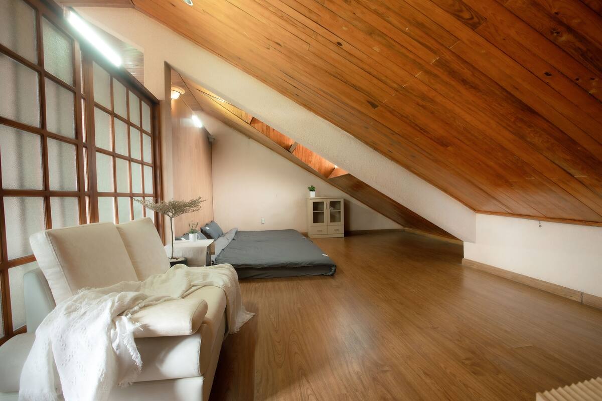 Quarto sótão com pé-direito alto, soalho em madeira, cama, sofá e um armário.