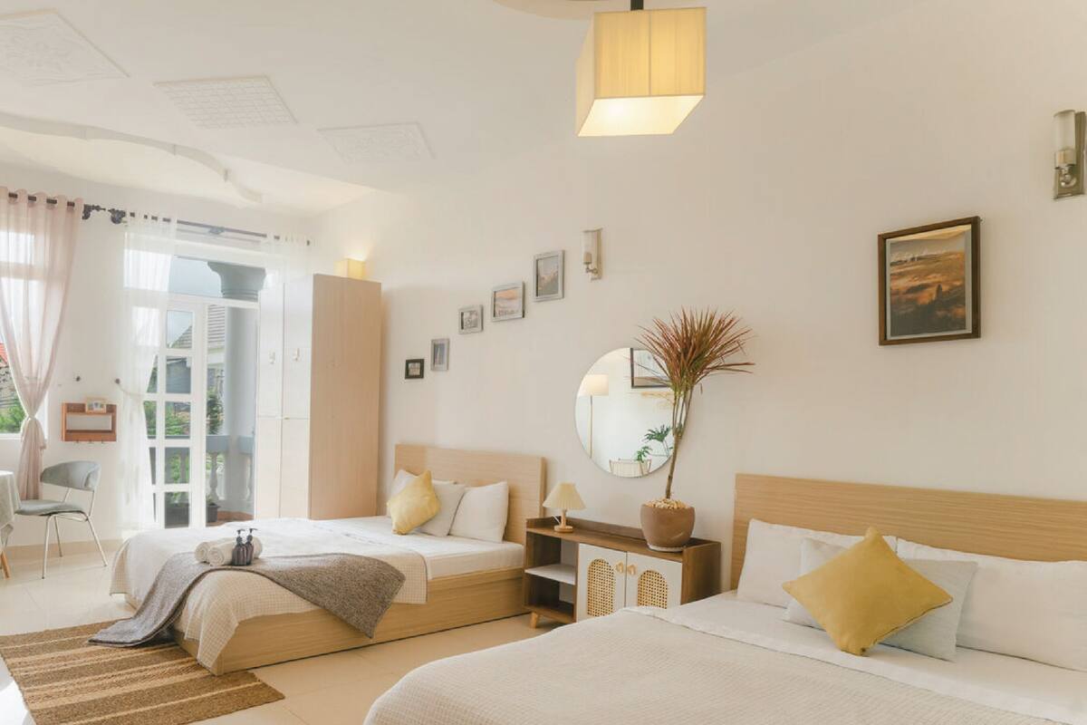 Quarto de hotel com duas camas, um espaço de estar com mesa e cadeira, uma planta em vaso e vista para o exterior.
