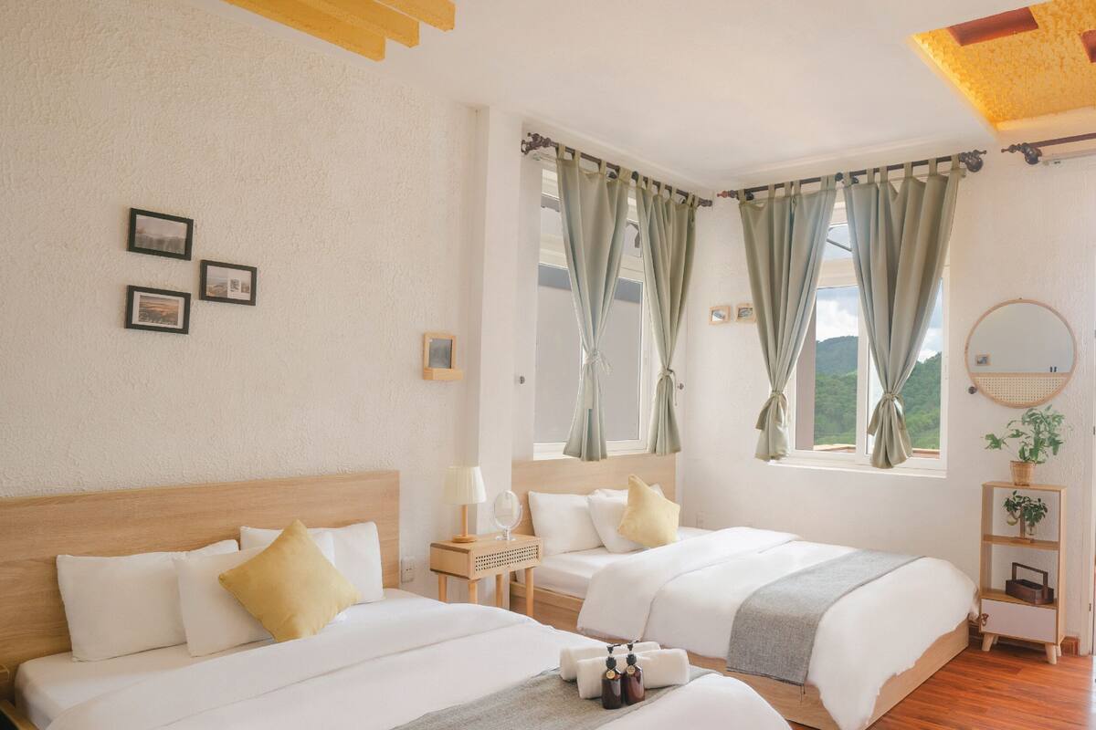 Quarto de hotel com duas camas, cabeceira de madeira, mesa de cabeceira com abajur, um pequeno espelho e uma janela com cortinas.