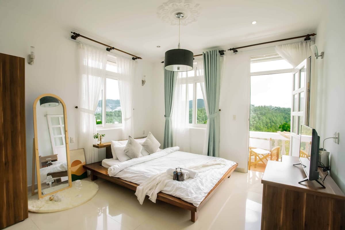 Quarto espaçoso com uma cama grande, mesa de cabeceira de madeira, espelho, escrivaninha com um computador e varanda com vista para o verde.