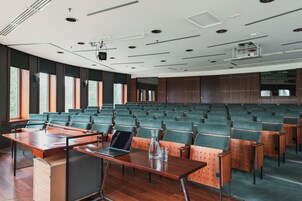 Uma sala de conferências com fileiras de cadeiras verdes, uma mesa de madeira com um laptop e um projetor no teto.